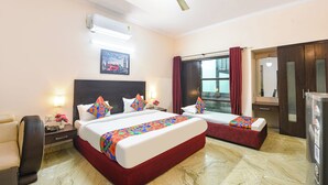 Quarto de hotel com duas camas, ventoinha no teto, aparelho de ar condicionado, mesa de cabeceira com abajur, janela com cortinas, espelho e guarda-roupa de madeira.