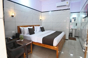 Quarto de hotel com cama, mesa de cabeceira, cadeira, planta em vaso e banheiro com espelho e pia.