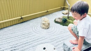 Criança sentada em superfície de pedra num jardim zen, com cerca de bambu ao fundo.