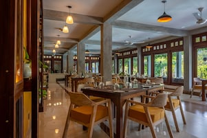 Uma sala de jantar espaçosa com mesas e cadeiras de madeira, janelas amplas e ventiladores de teto.
