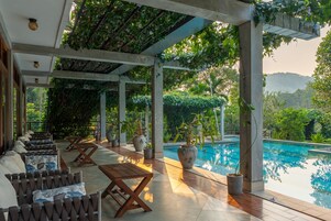 Uma área de estar coberta ao ar livre com piscina, móveis de madeira e plantas em vasos.