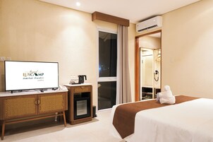 Quarto de hotel com uma cama, uma televisão sobre um armário de madeira, uma janela com cortinas e um aparelho de ar condicionado.