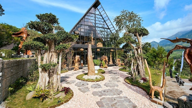 Um edifício moderno com estrutura em forma de pirâmide de vidro, cercado por um jardim com esculturas e árvores.