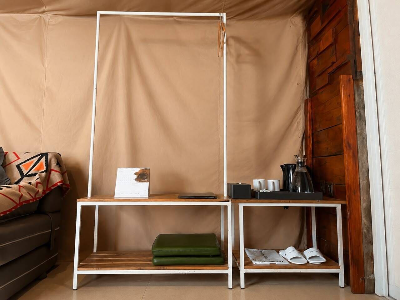 Quarto com divisória branca, um banco verde, uma mesa pequena com um caderno, uma cafeteira e chinelos.