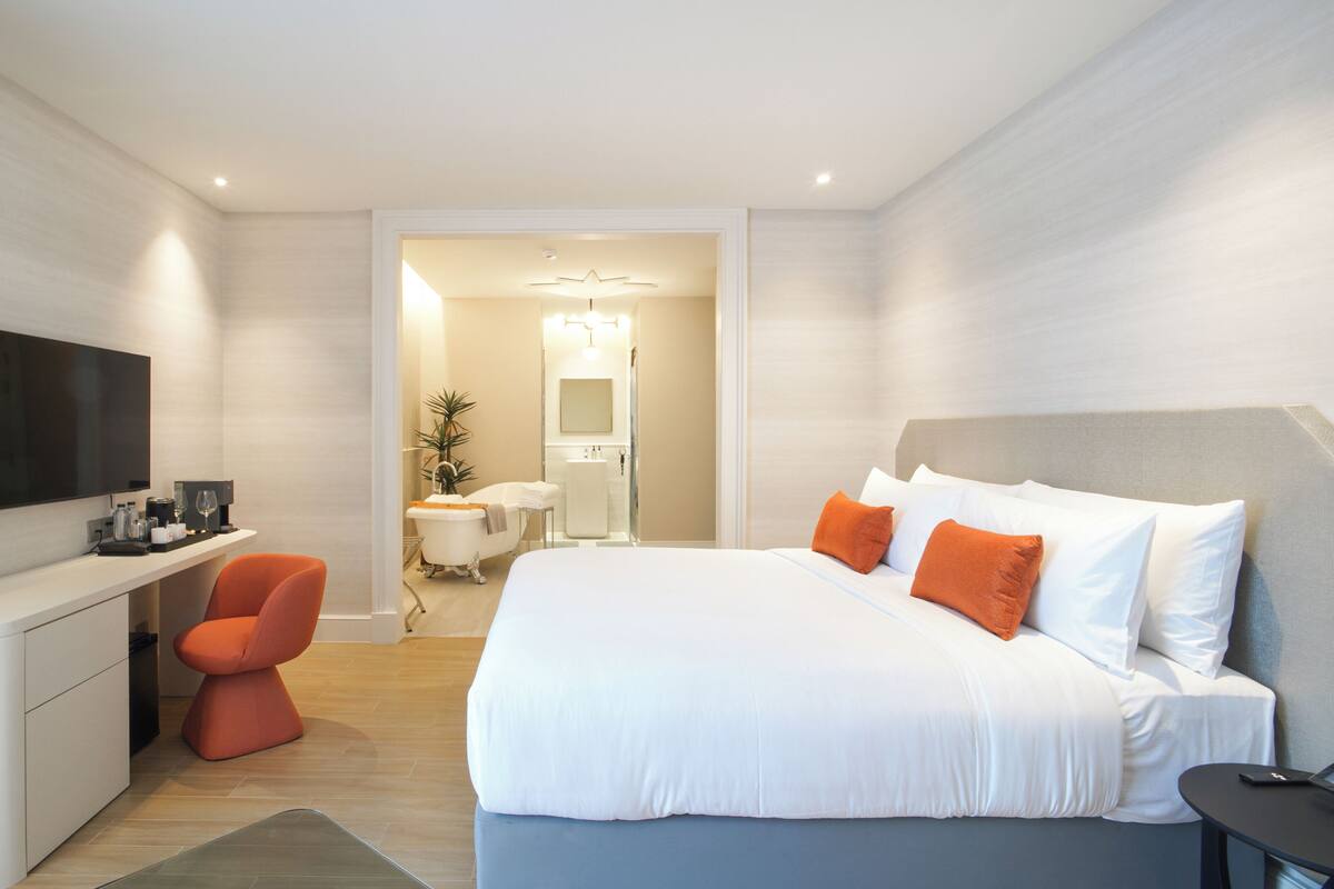Quarto de hotel com cama, escrivaninha, cadeira, televisão e banheiro visível por uma porta aberta.