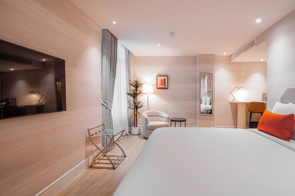 Quarto de hotel moderno com uma cama grande, uma escrivaninha, uma cadeira, uma planta em vaso e um espelho na parede.