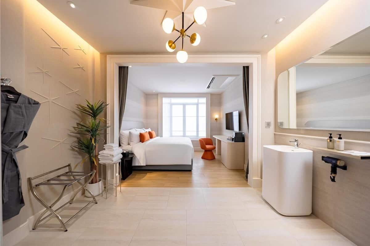 Quarto de hotel moderno com cama, escrivaninha, cadeira, espelho grande e banheiro com pia e vaso sanitário.