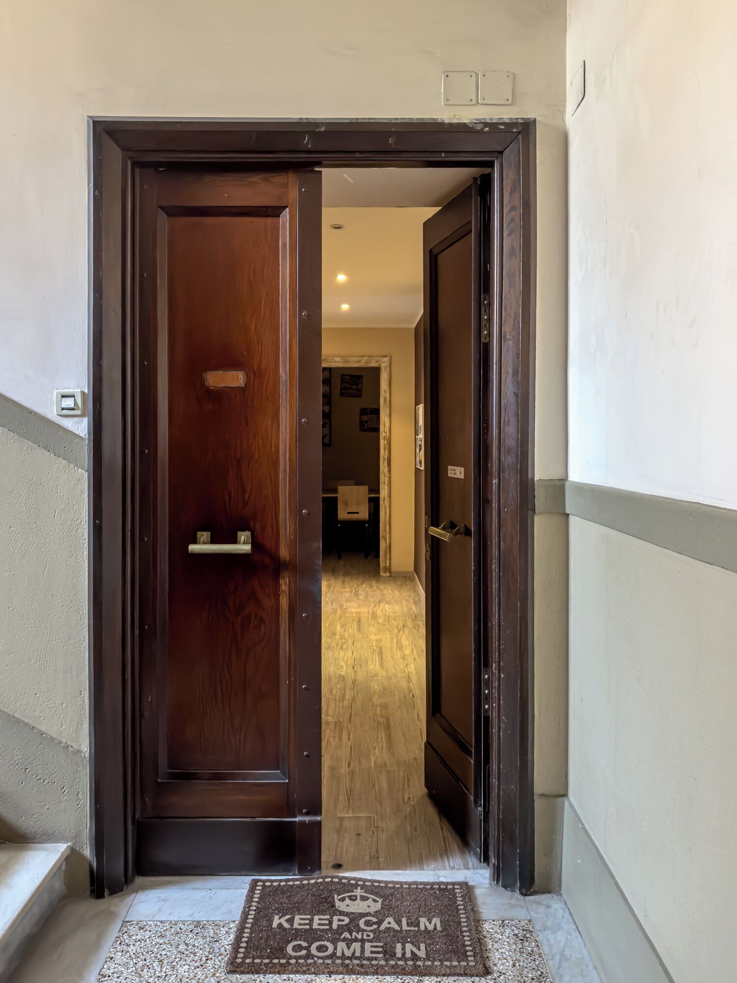 Um corredor com porta de madeira, tapete de entrada e piso claro.