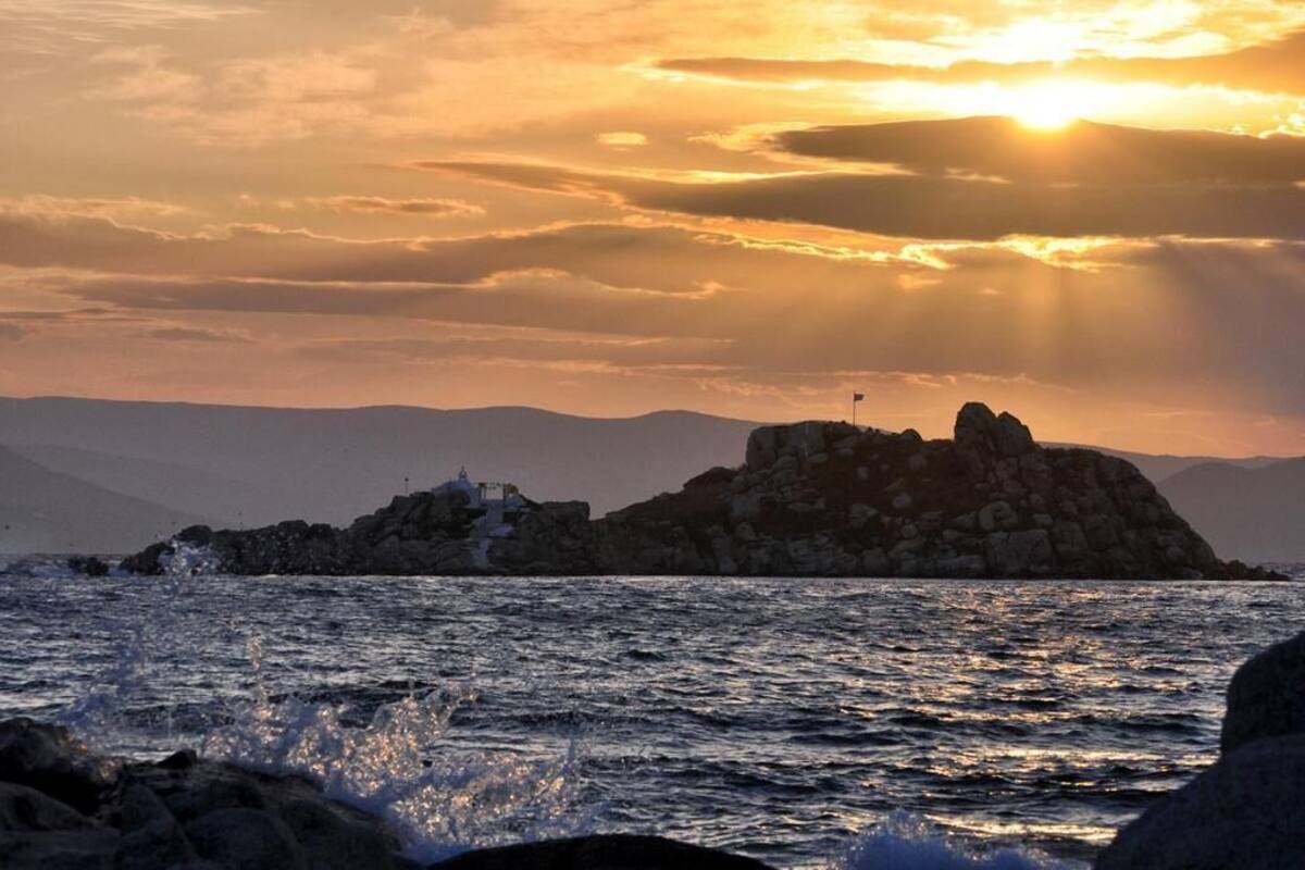 Cena costeira com um afloramento rochoso, ondas e uma cordilheira distante.