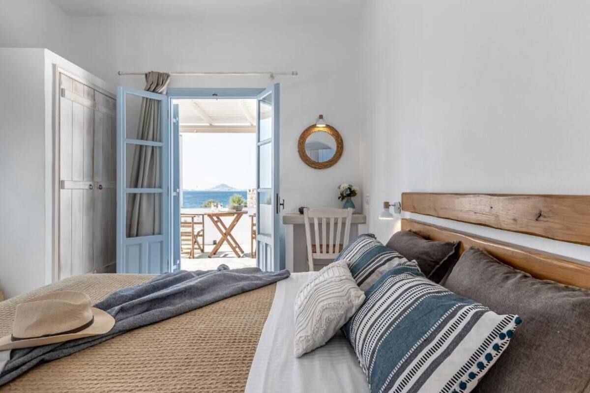 Quarto com cama, vista para o mar através de uma porta aberta, cabeceira de madeira e uma pequena mesa com cadeira.