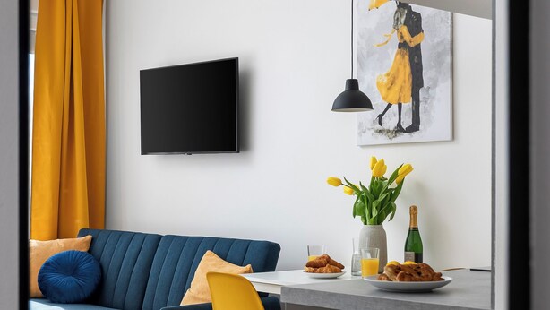 Uma sala de estar moderna com um sofá, uma poltrona amarela, uma televisão de tela plana, um quadro, um vaso com flores e uma garrafa de champanhe.