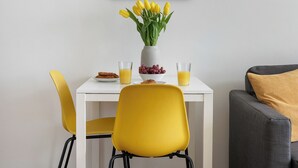 Uma sala de jantar com uma mesa branca, cadeiras amarelas, um vaso com flores amarelas e uma obra de arte emoldurada acima.