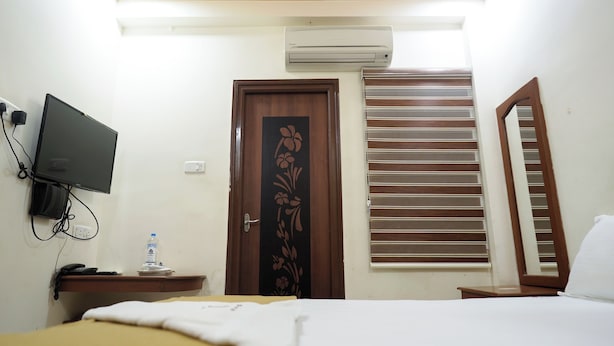 Quarto de hotel com uma cama, televisão fixada na parede, aparelho de ar condicionado, porta com desenho floral e janela com persianas.