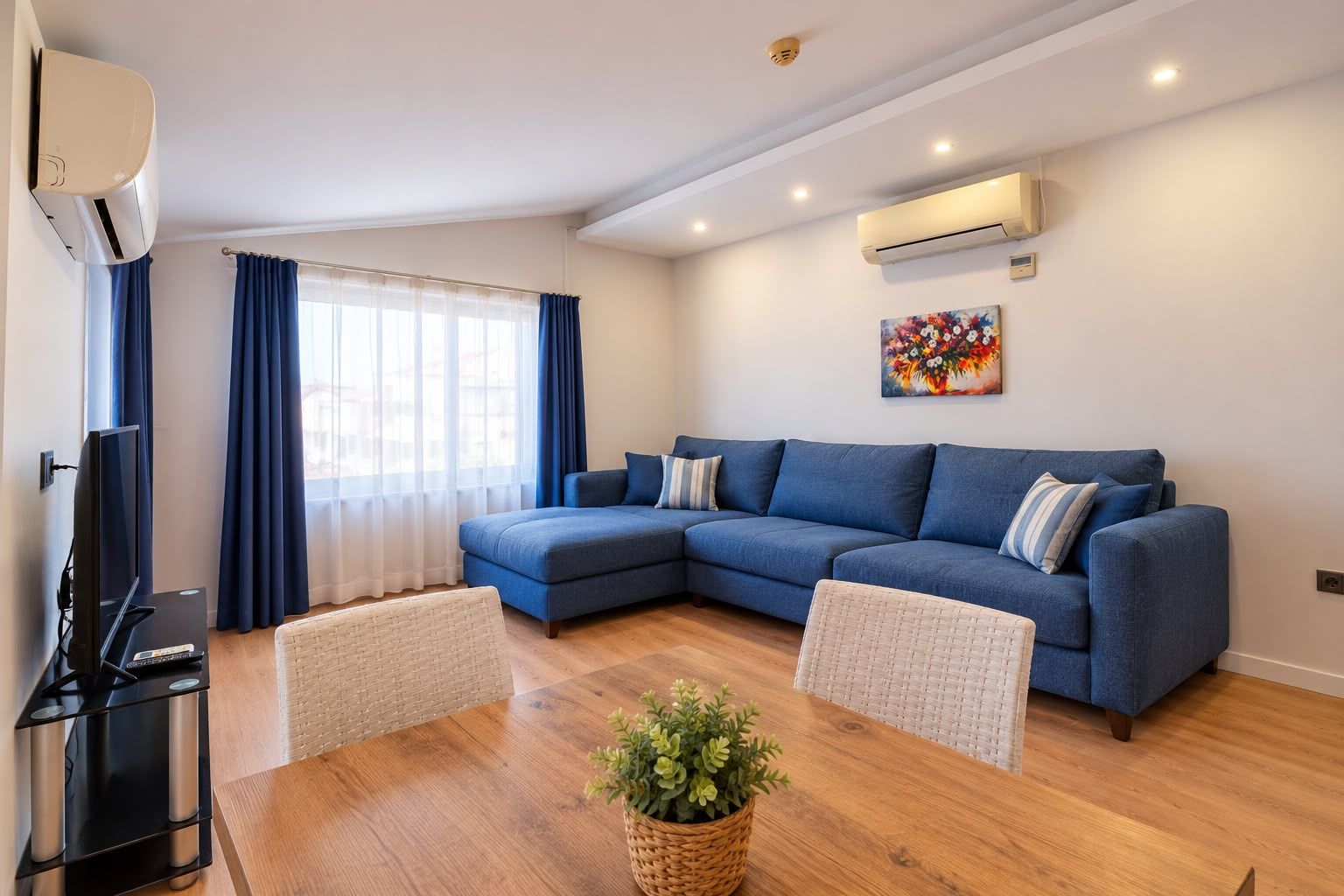Uma sala de estar moderna com um sofá seccional, uma TV de tela plana, uma mesa de jantar de madeira e uma planta em vaso.