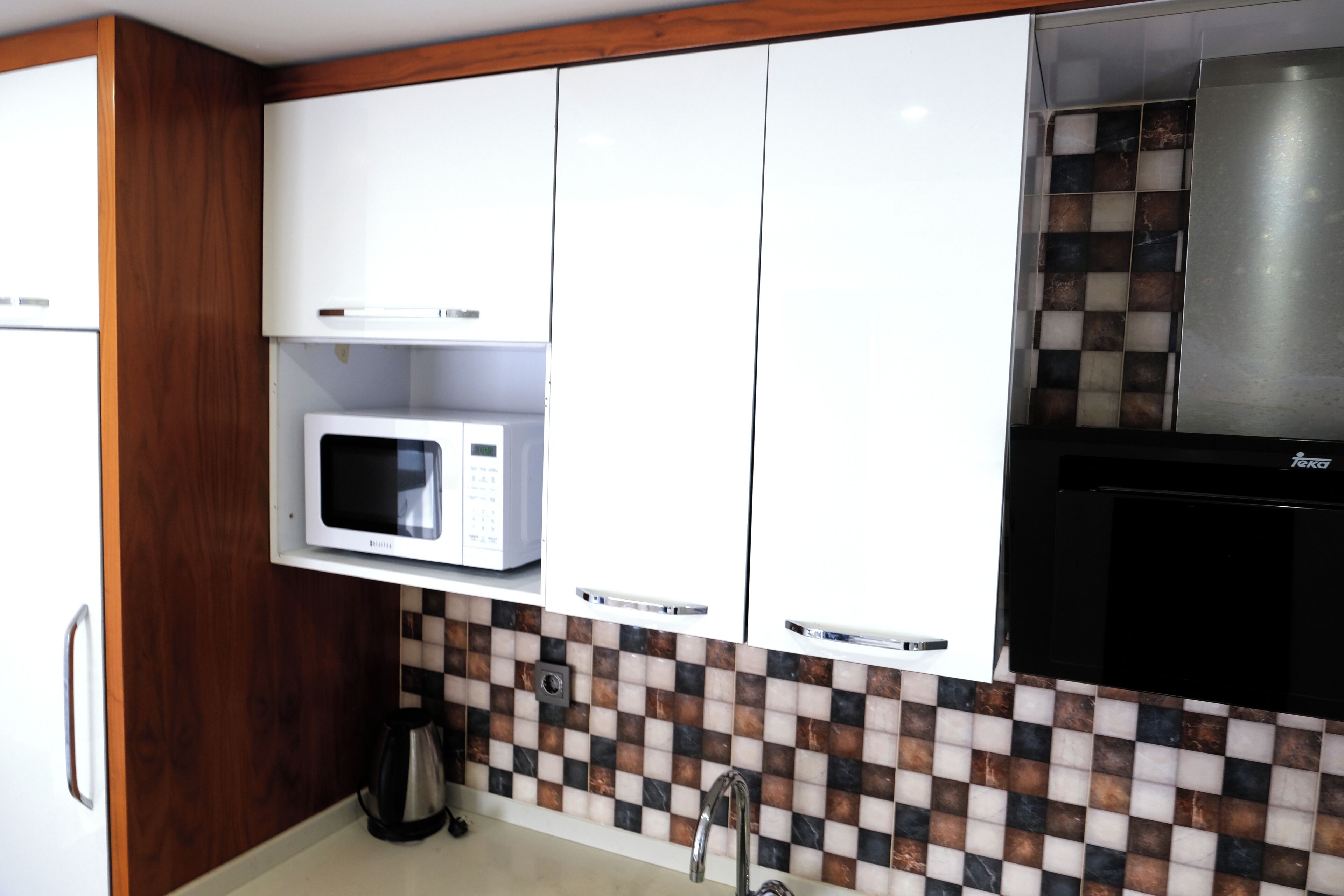 Cozinha com armários brancos, micro-ondas, chaleira e painel de azulejos.