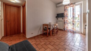 Um quarto com piso de azulejos, uma mesa de jantar com cadeiras, uma televisão fixada na parede e uma porta com moldura de madeira.