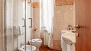 Uma casa de banho com cabine de chuveiro em vidro, sanita, lavatório e tapete branco no chão.