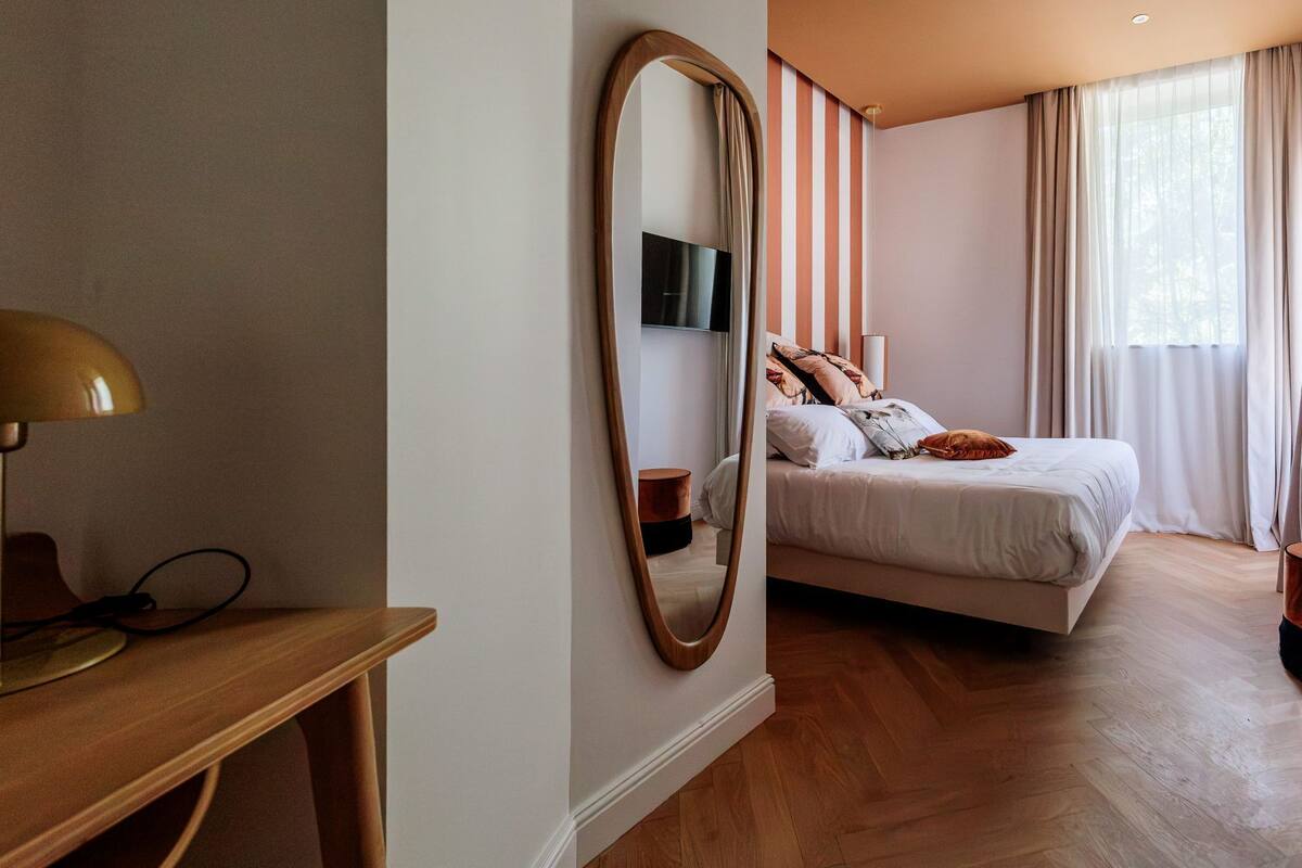 Um quarto com cama, escrivaninha com luminária, espelho e televisão na parede.