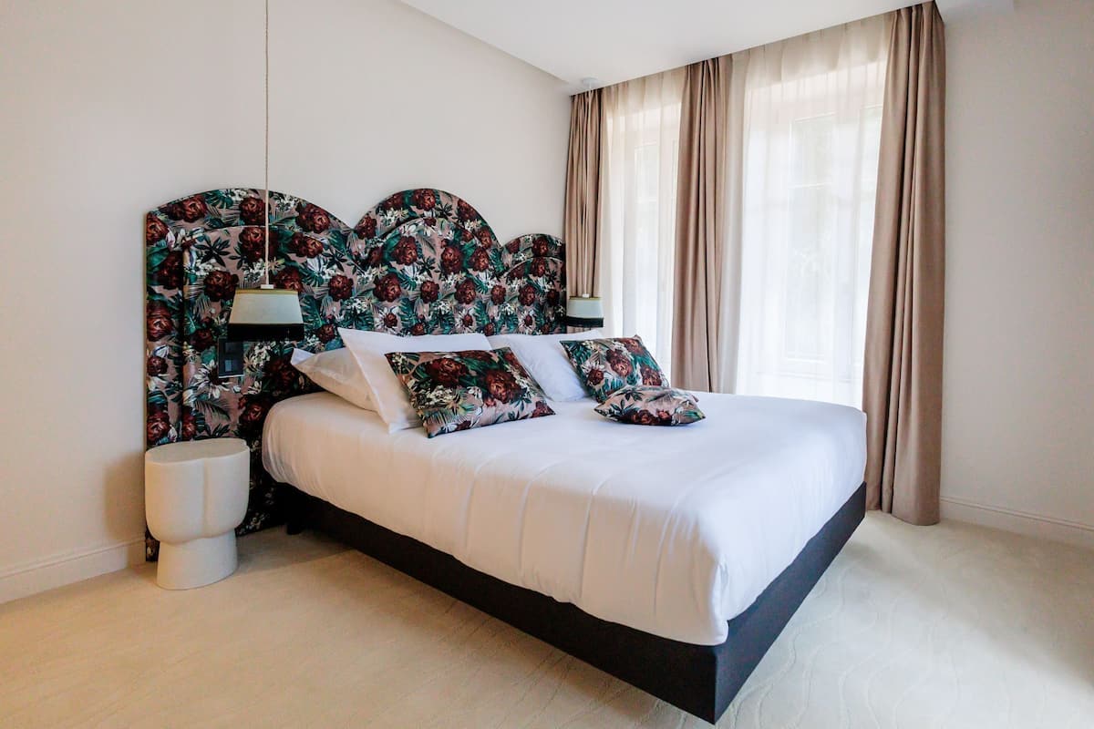 Uma cama bem arrumada com almofadas estampadas e cabeceira, uma luminária de mesa e uma janela com cortinas.