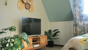 Quarto com televisão de tela plana sobre suporte de madeira, planta em vaso e cama com colcha florida.