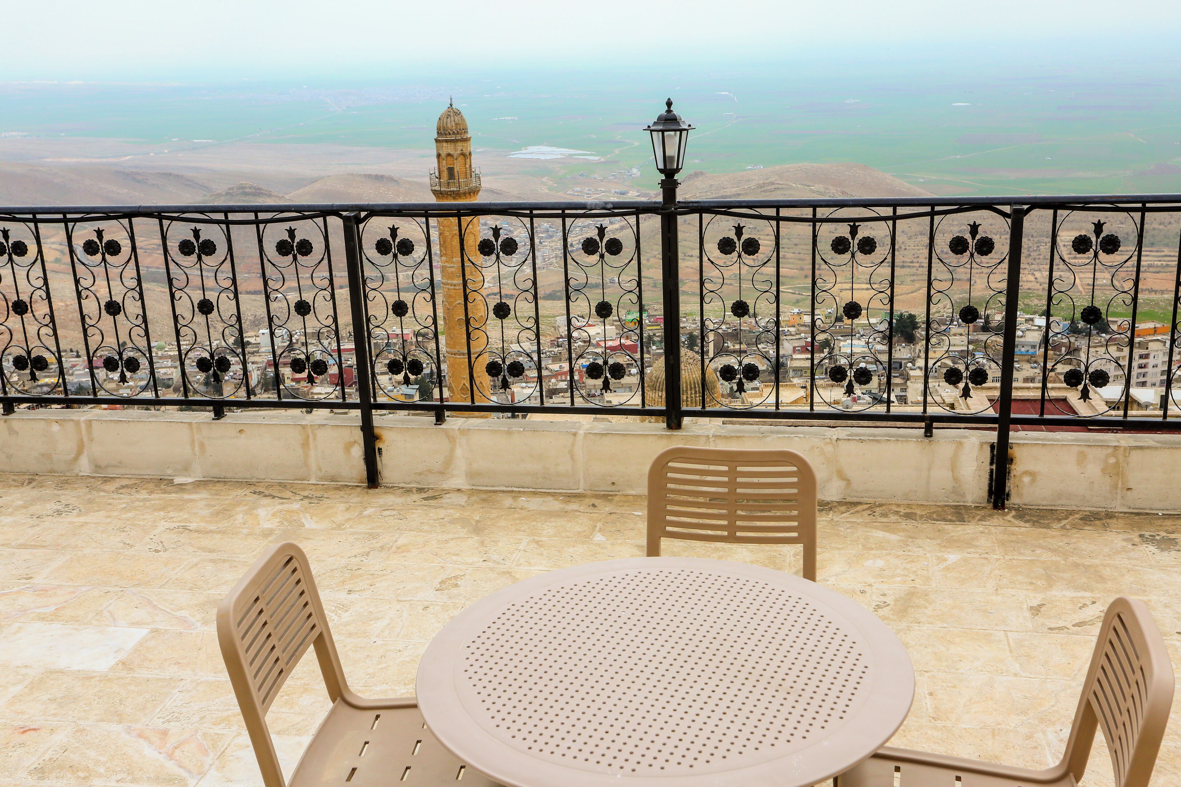 Uma varanda com mesa e cadeiras, um grade decorativo e vista para uma paisagem distante com uma torre.