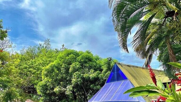 Uma área de piscina com um pavilhão de telhado azul, cercada por vegetação exuberante e um céu claro.