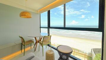 Quarto com uma grande janela com vista para a praia, uma mesa redonda, duas cadeiras, um sofá e um lustre.