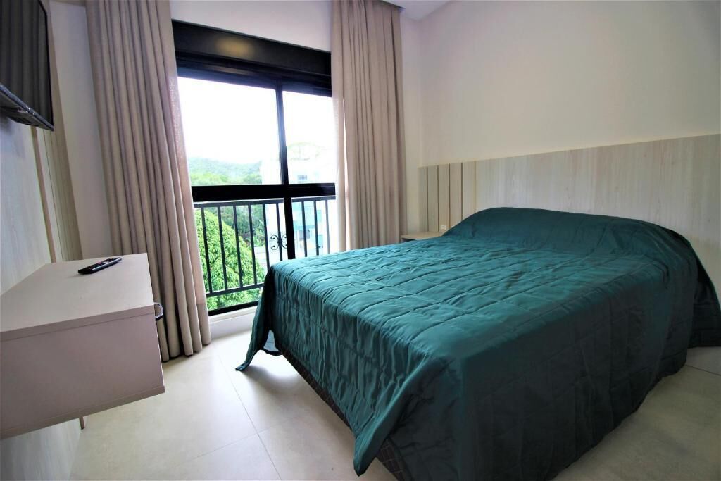 Quarto com cama, mesa de cabeceira, janela com cortinas e varanda.