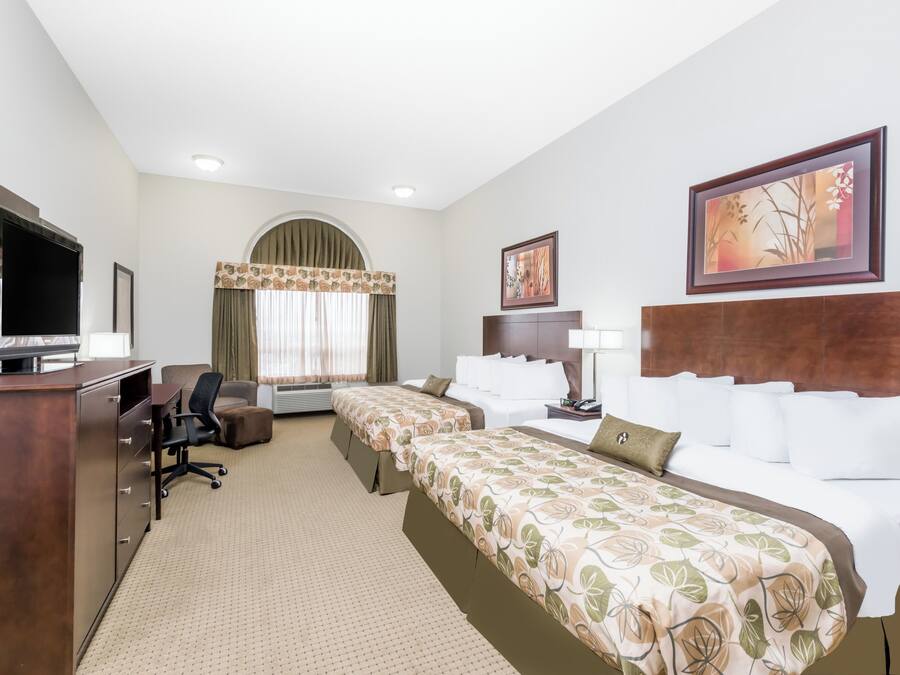 Quarto de hotel com televisão, secretária, duas camas, uma cadeira, luminária de mesa, quadro emoldurado e janela com cortinas.