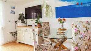 Uma área de jantar com mesa de vidro, cadeiras florais, um armário branco, uma TV de tela plana e uma pintura de veleiros na parede.