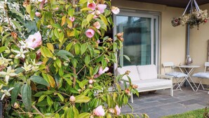 Um jardim com plantas floridas, uma área de estar externa com assentos brancos e uma porta de vidro que dá para o interior.