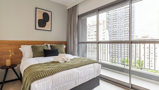Quarto de hotel moderno com uma cama grande, uma mesinha de cabeceira com um vaso, um quadro na parede e vista para edifícios altos.
