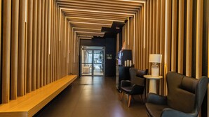 Um hall de hotel moderno com parede de ripas de madeira, balcão de recepção, área de estar e entrada por porta de vidro.