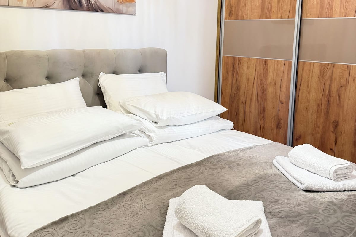 Cama bem arrumada com lençóis brancos, colcha cinza estampada e guarda-roupa de madeira com portas de correr.