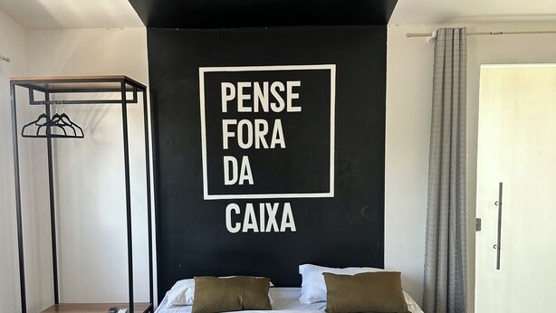 Quarto de hotel com cama, prateleira na parede, guarda-roupa e um aviso com texto.