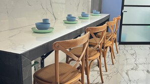 Uma área de refeições com piso de mármore, uma mesa comprida com tigelas azuis e verdes, e cadeiras de vime.