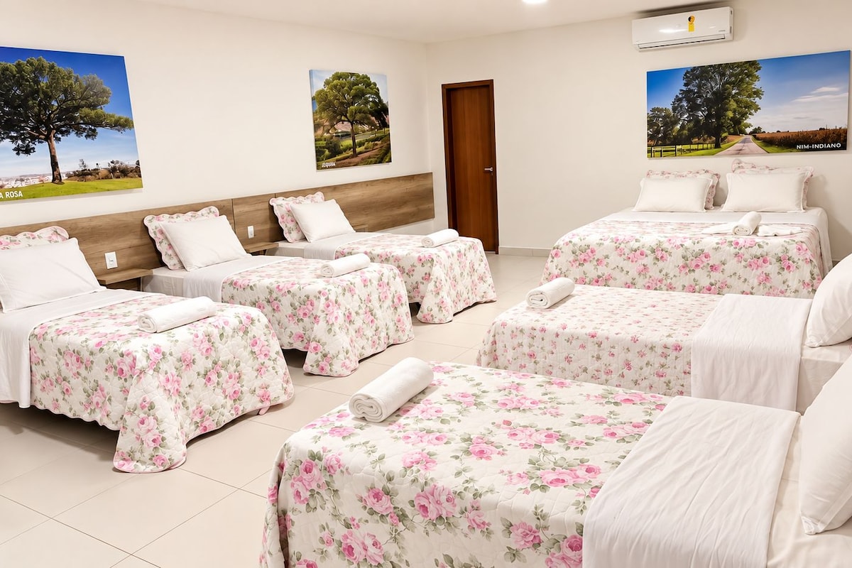 Quarto de hotel com quatro camas, cada uma com roupa de cama floral, cabeceira de madeira e uma janela grande.