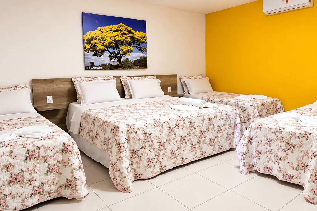 Quarto de hotel com três camas, cada uma com colcha florida, mesa de cabeceira, ar-condicionado fixado na parede e quadro com uma árvore amarela.