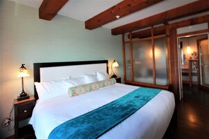 Quarto de hotel com uma cama, duas luminárias de cabeceira, teto de madeira e uma porta deslizante para um banheiro.