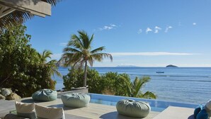 Piscina infinita com espreguiçadeiras e almofadas, com vista para o mar azul cristalino e uma ilha ao longe.
