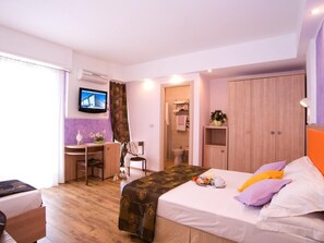 Quarto com cama, escrivaninha, cadeira, televisão e banheiro visível por uma porta aberta.