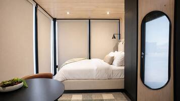 Quarto de hotel moderno com uma cama, uma mesinha com uma planta, vista pela janela e um espelho redondo.