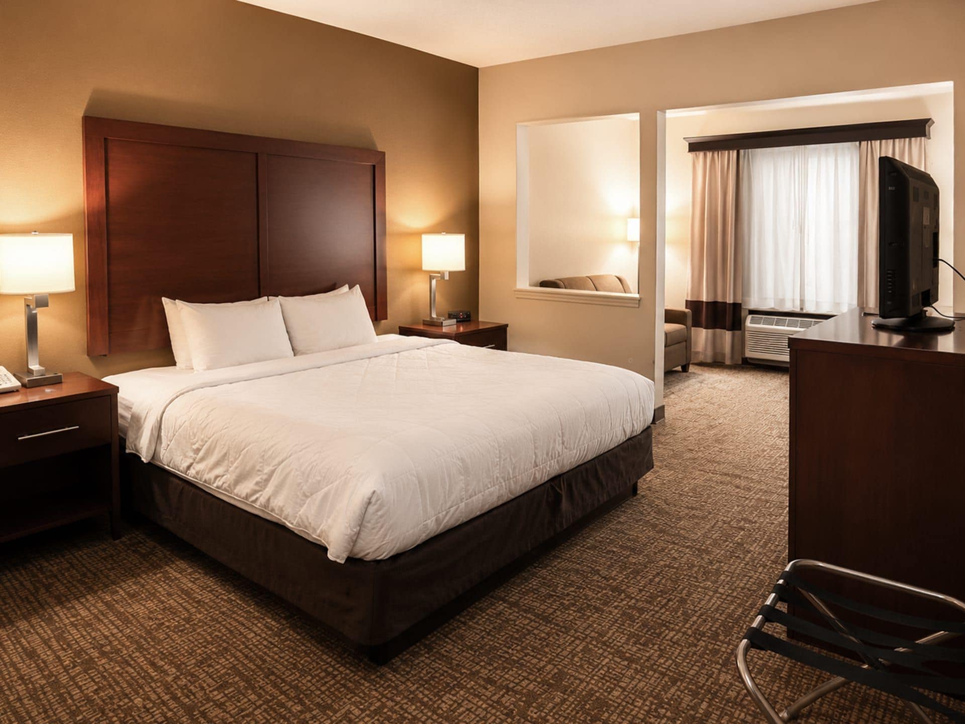 Quarto de hotel com uma cama grande, mesas de cabeceira com luminárias, uma escrivaninha com computador, uma cadeira e vista para o exterior pela janela.