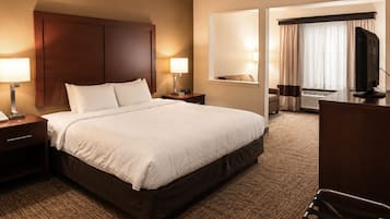 Quarto de hotel com uma cama grande, mesas de cabeceira com luminárias, uma escrivaninha com computador, uma cadeira e vista para o exterior pela janela.
