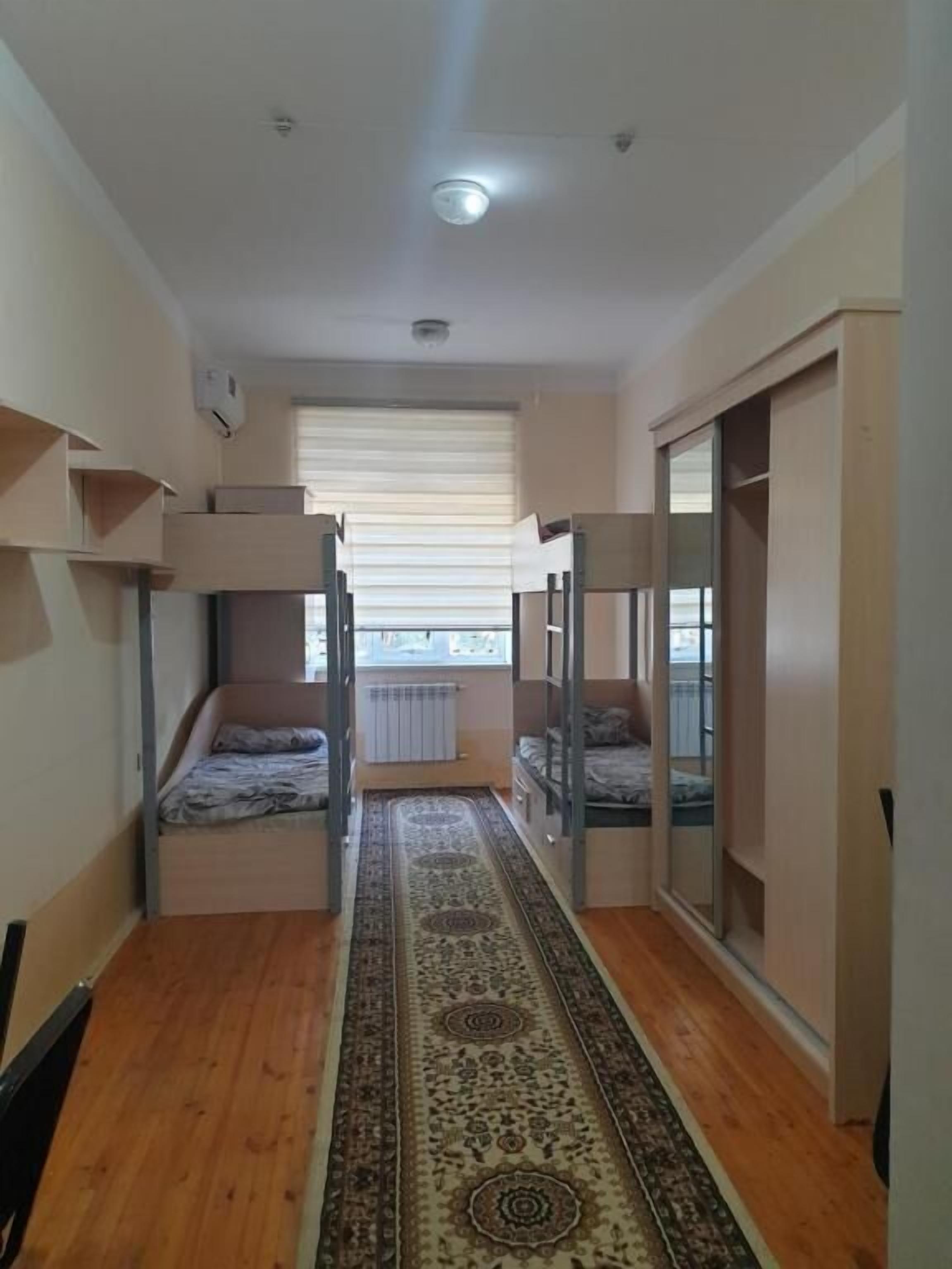Quarto pequeno com beliches, radiador, janela com persianas e tapete estampado.