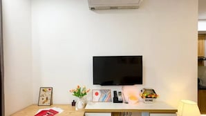 Quarto com televisão de tela plana na parede, escrivaninha com luminária, prateleira pequena e quadro emoldurado.
