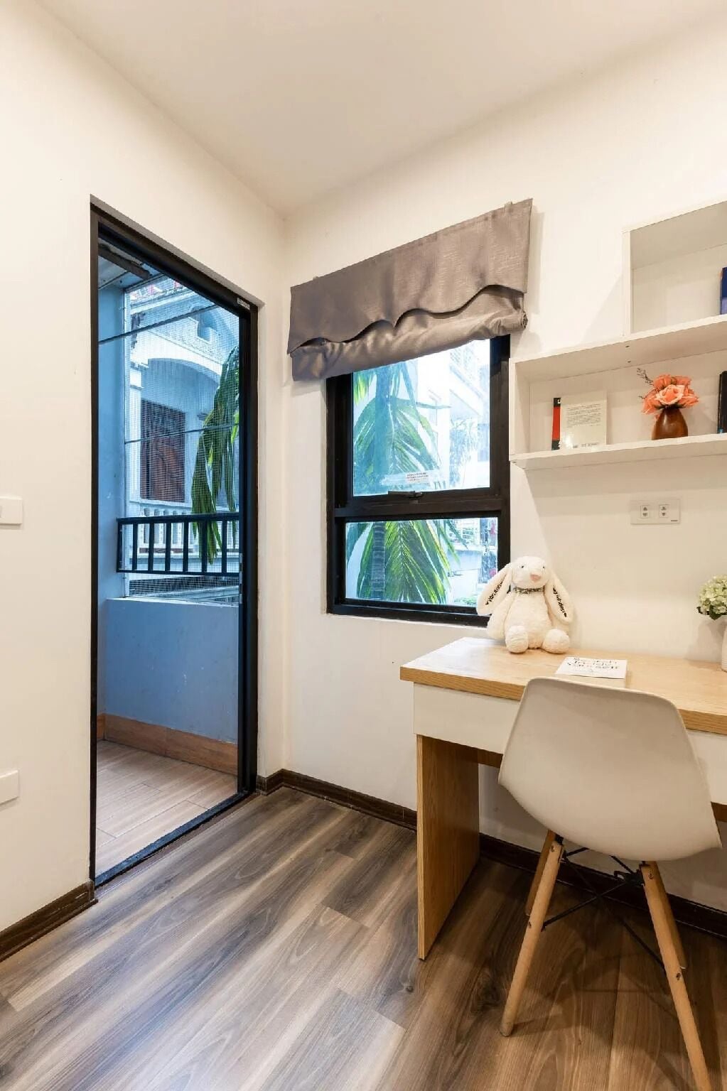 Quarto com escrivaninha, cadeira e janela com vista para uma varanda e área verde.