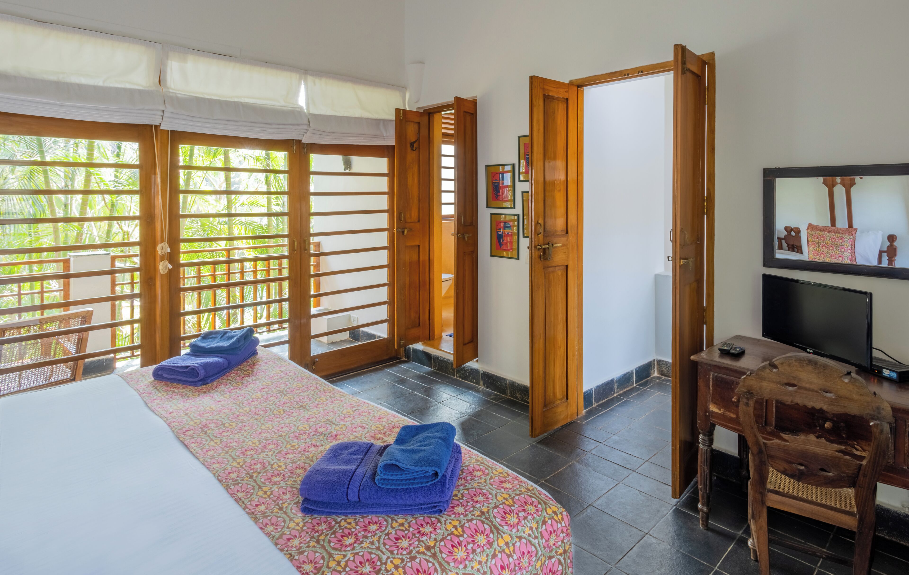 Quarto de hotel com cama, escrivaninha com televisão e varanda com vista para palmeiras.