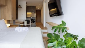 Quarto de hotel moderno com cama, televisão na parede, cozinha compacta ao fundo e uma planta grande.