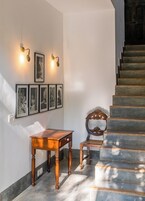 Uma escadaria com uma cadeira de madeira e uma mesa, uma parede com quadros emoldurados, e um luminário.